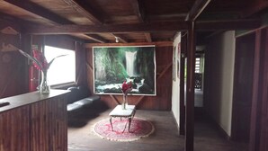 Quarto com teto de madeira, uma grande pintura de uma cachoeira, uma mesa pequena com um vaso de flores e um sofá de couro preto.