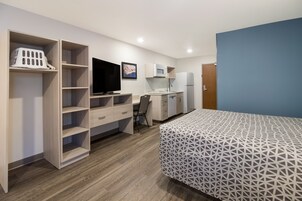 Quarto compacto com uma cama, escrivaninha com computador, televisão e cozinha compacta.