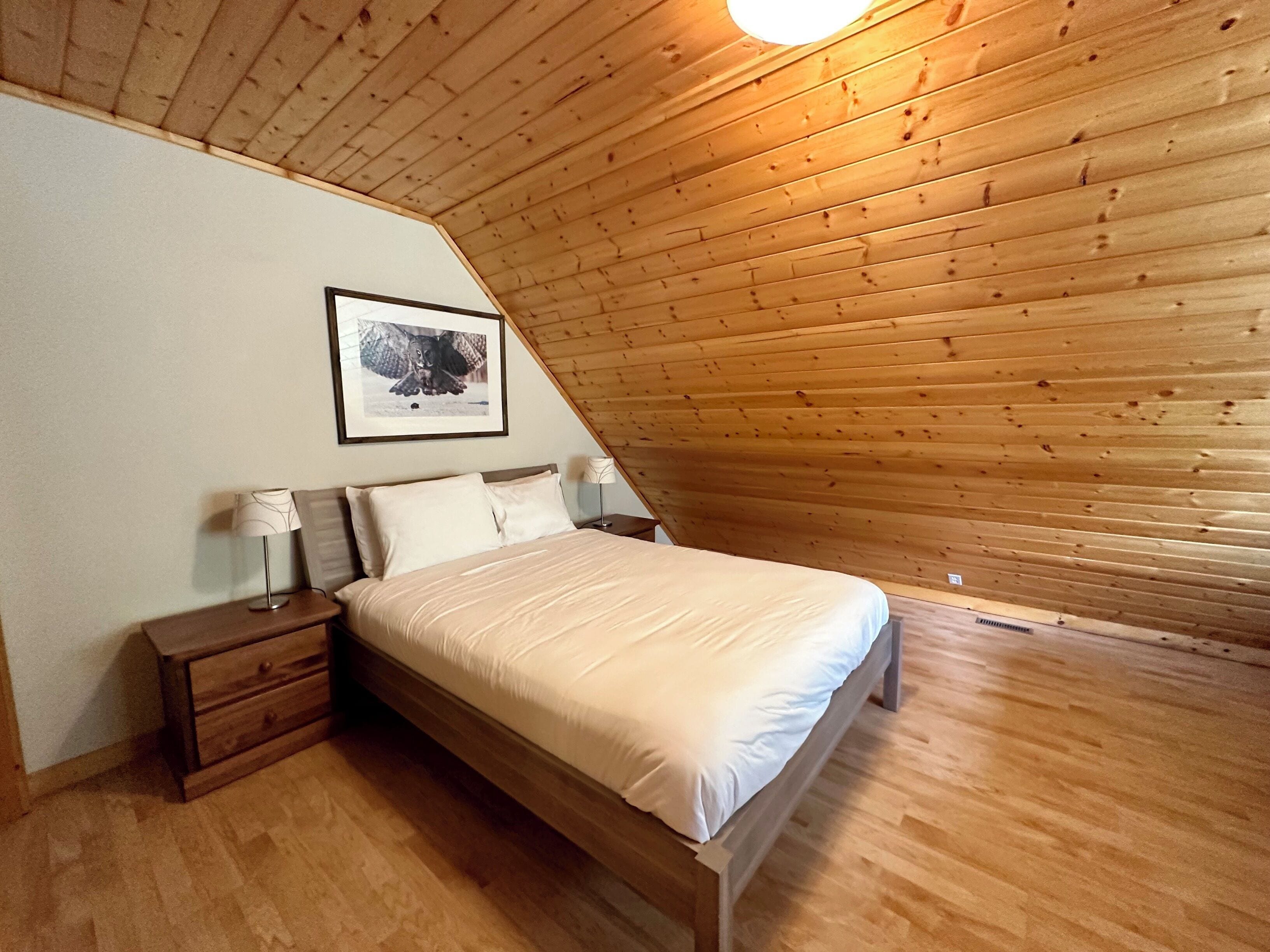 Quarto com teto de madeira, cama com roupa de cama branca, mesa de cabeceira, abajur e um quadro com uma coruja.