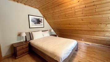 Quarto com teto de madeira, cama com roupa de cama branca, mesa de cabeceira, abajur e um quadro com uma coruja.