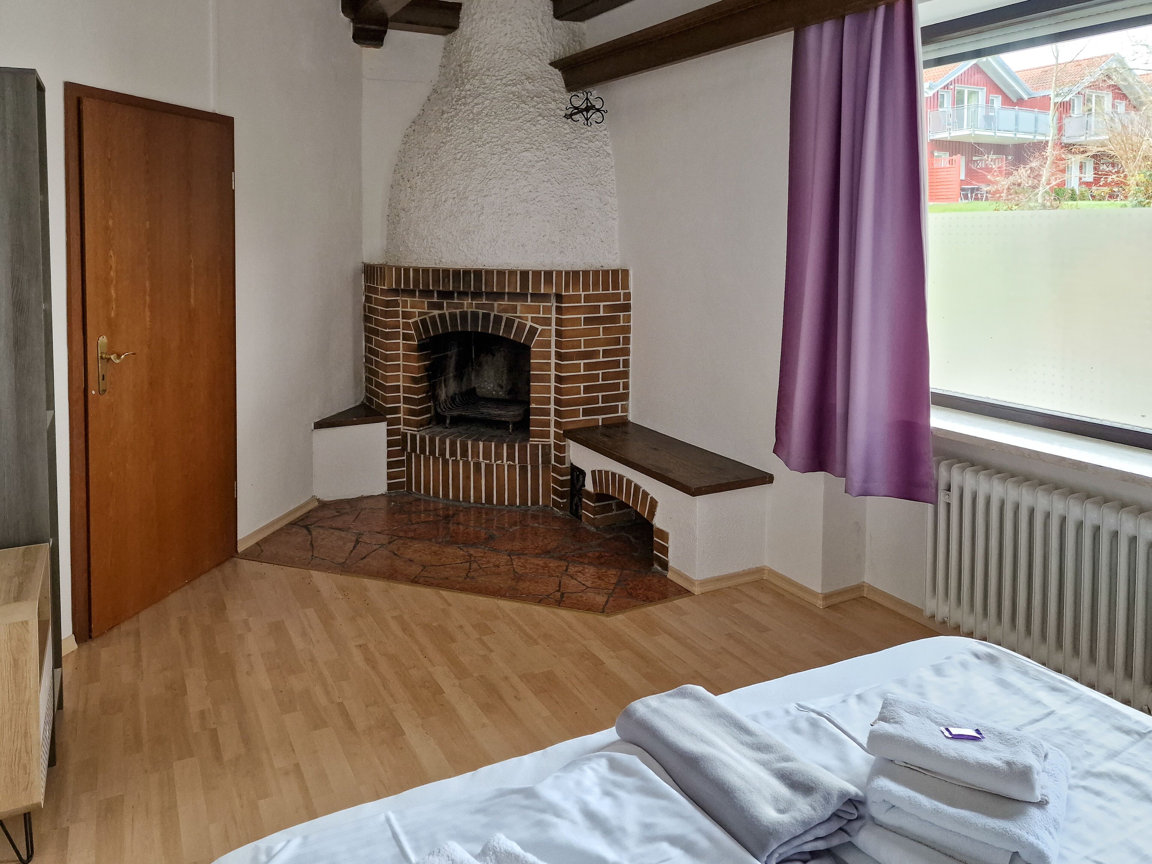 Quarto com lareira de tijolos, piso de madeira, cama com roupa de cama branca, janela com cortinas roxas e vista para um prédio vermelho lá fora.