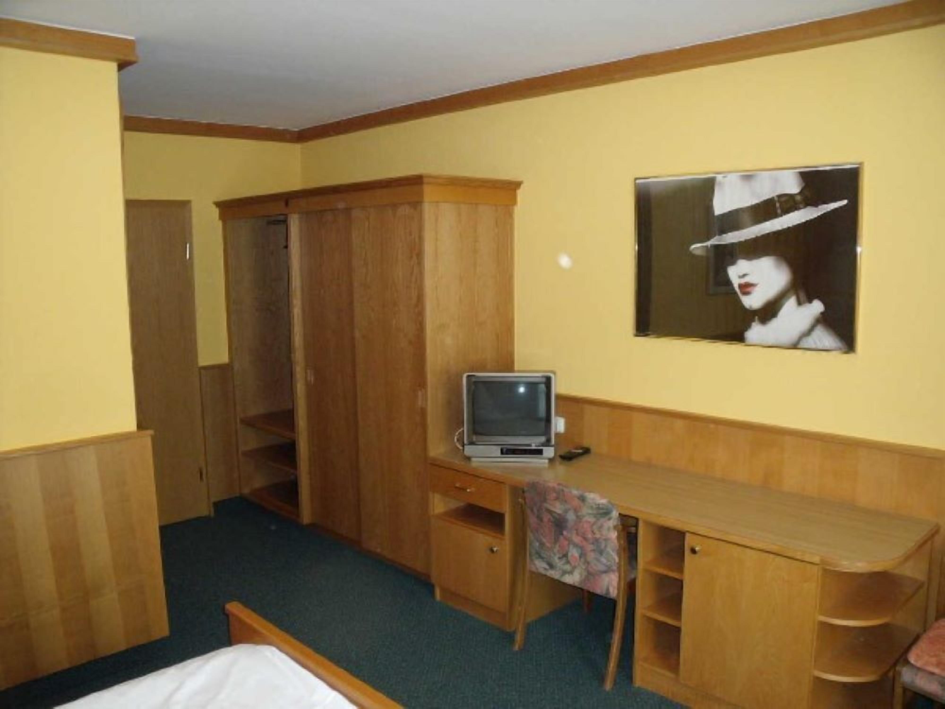 Quarto de hotel compacto com uma secretária de madeira, televisão, guarda-roupa e uma cama.