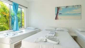 Quarto de hotel com duas camas, uma janela com cortinas, um quadro de uma praia e vista para o verde lá fora.