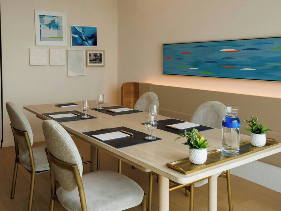Uma sala de conferências com uma mesa comprida, quatro cadeiras, um copo de água, um vaso e quadros na parede.