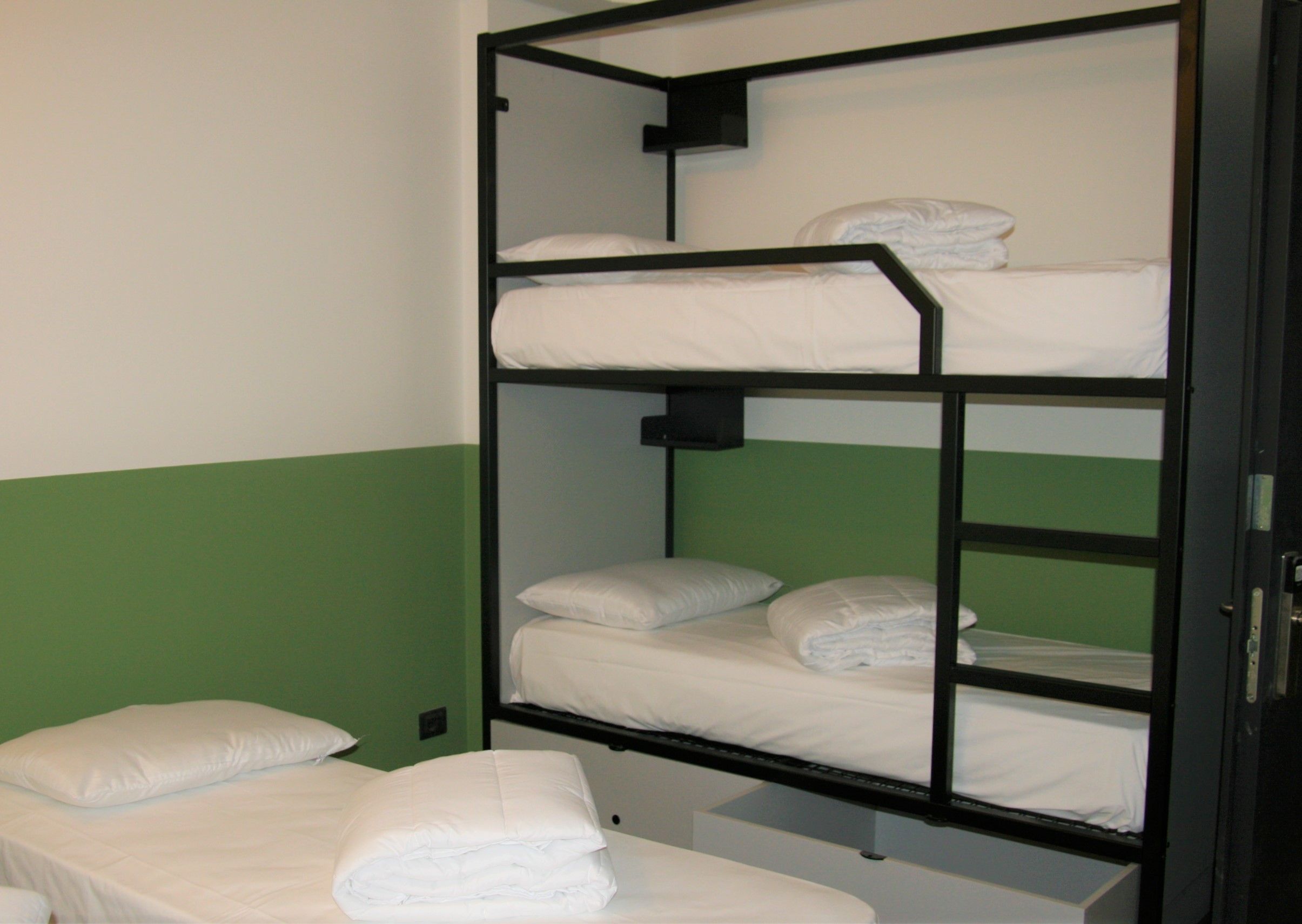 Uma cama beliche com roupa de cama e almofadas brancas, uma parede verde e uma porta à direita.