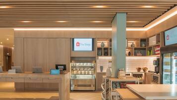 Um saguão de hotel com balcão de recepção, um painel digital para 'The Shop', um minibar e uma área de estar.