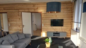 Sala de estar com um sofá cinza, uma mesa de centro preta com uma planta, uma TV fixada na parede e uma parede de madeira.