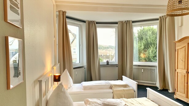 Quarto de hotel com duas camas, um radiador, um guarda-roupa de madeira e vista para o verde lá fora.