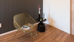 Quarto moderno com piso de madeira escura, uma cadeira de fio dourada, uma pequena mesa preta com um vaso e incensos, e um relógio fixado na parede.