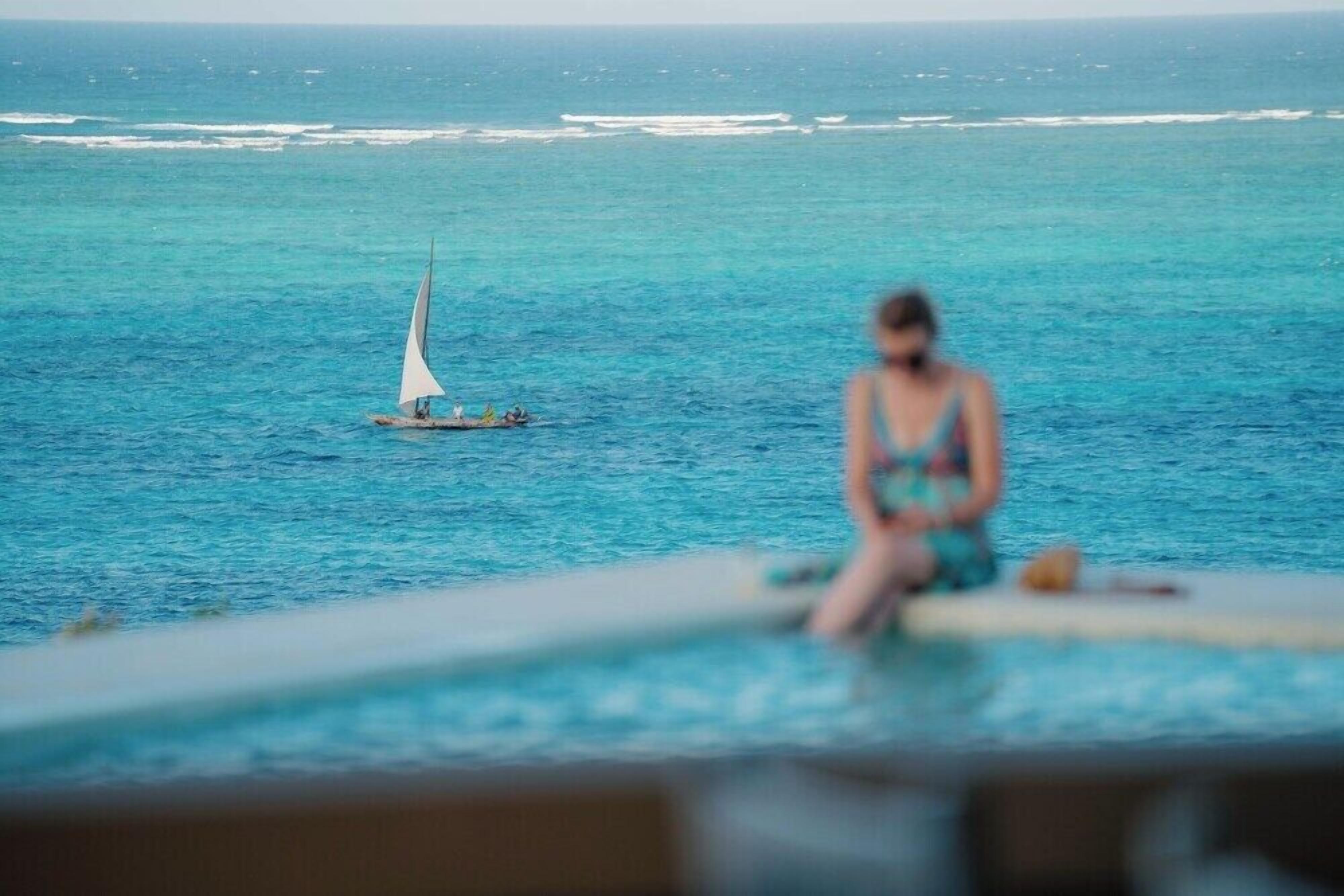Uma pessoa sentada à beira da piscina, com vista para o oceano e um veleiro ao longe.
