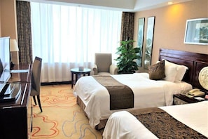 Quarto de hotel com duas camas, uma escrivaninha, uma cadeira, uma mesinha e uma planta grande.