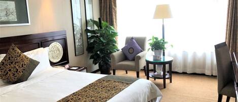 Quarto de hotel com uma cama grande, duas poltronas, uma mesinha e uma janela com cortinas.
