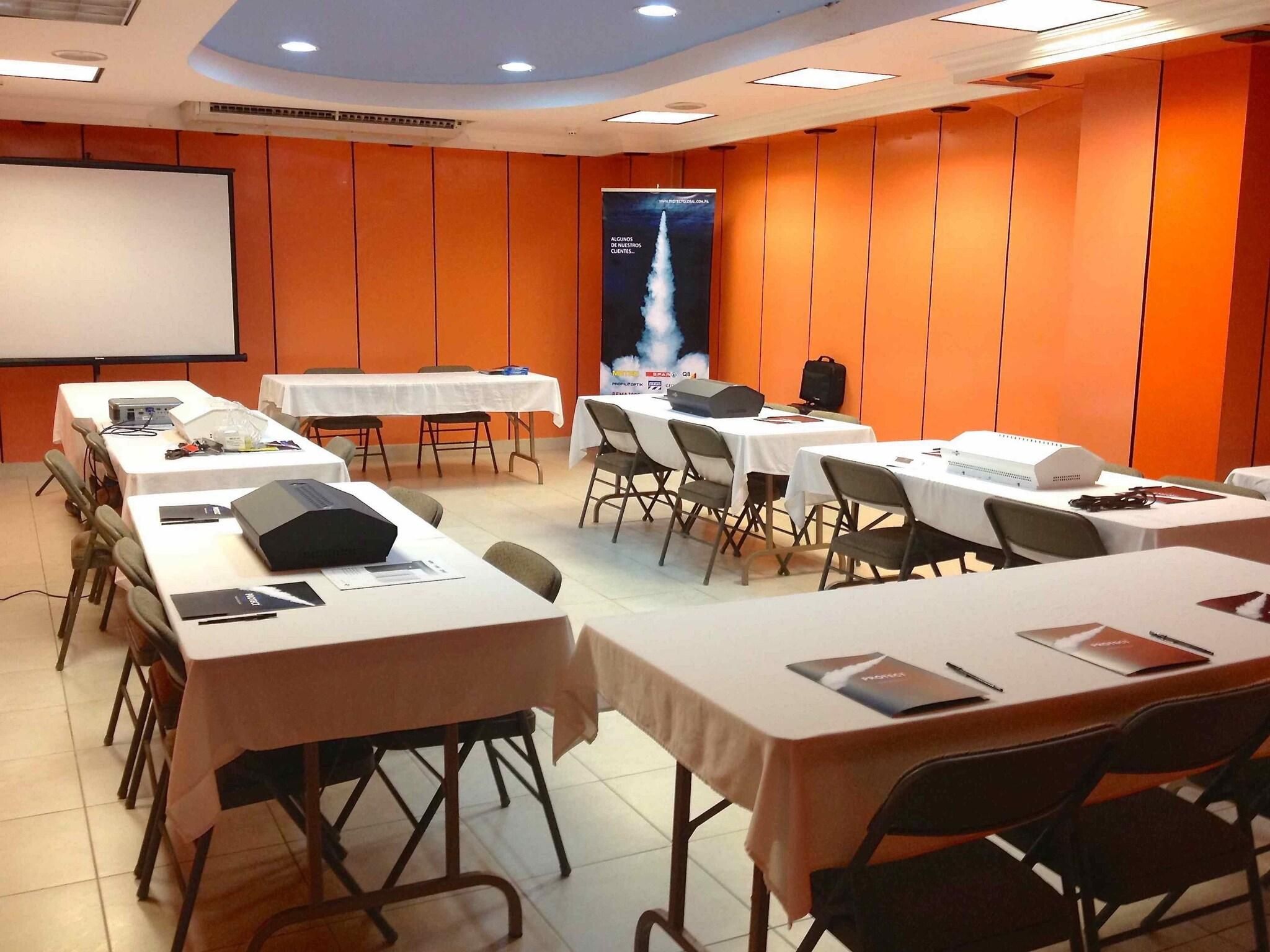 Sala de conferência com mesas, cadeiras, projetor e tela.
