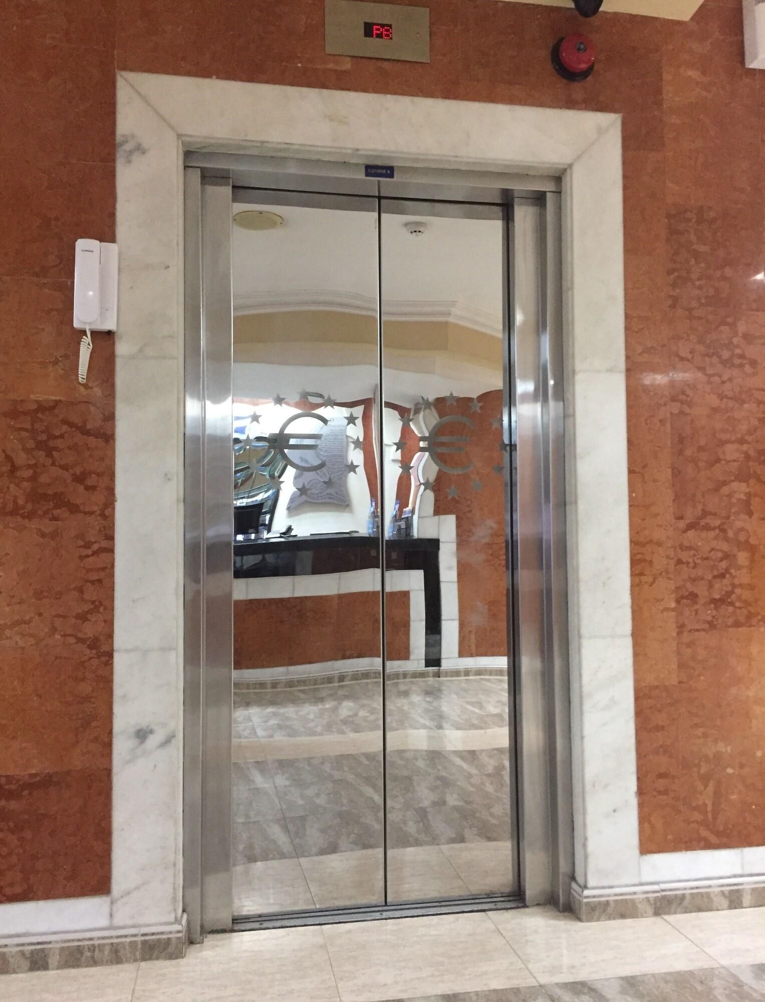 Porta de elevador dupla com um painel digital acima, um telefone e uma parede com padrão marmorizado.