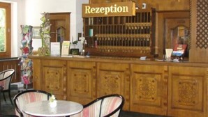 Uma área de recepção com balcão de madeira, uma placa com a inscrição "Rezeption" e uma janela com vista para o verde. Há cadeiras e uma mesa em frente ao balcão.