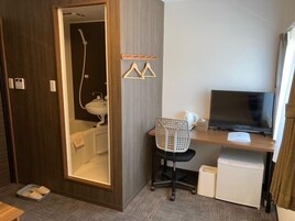 Quarto de hotel com escrivaninha de madeira, televisão, cadeira, pequena geladeira, pia e chuveiro.