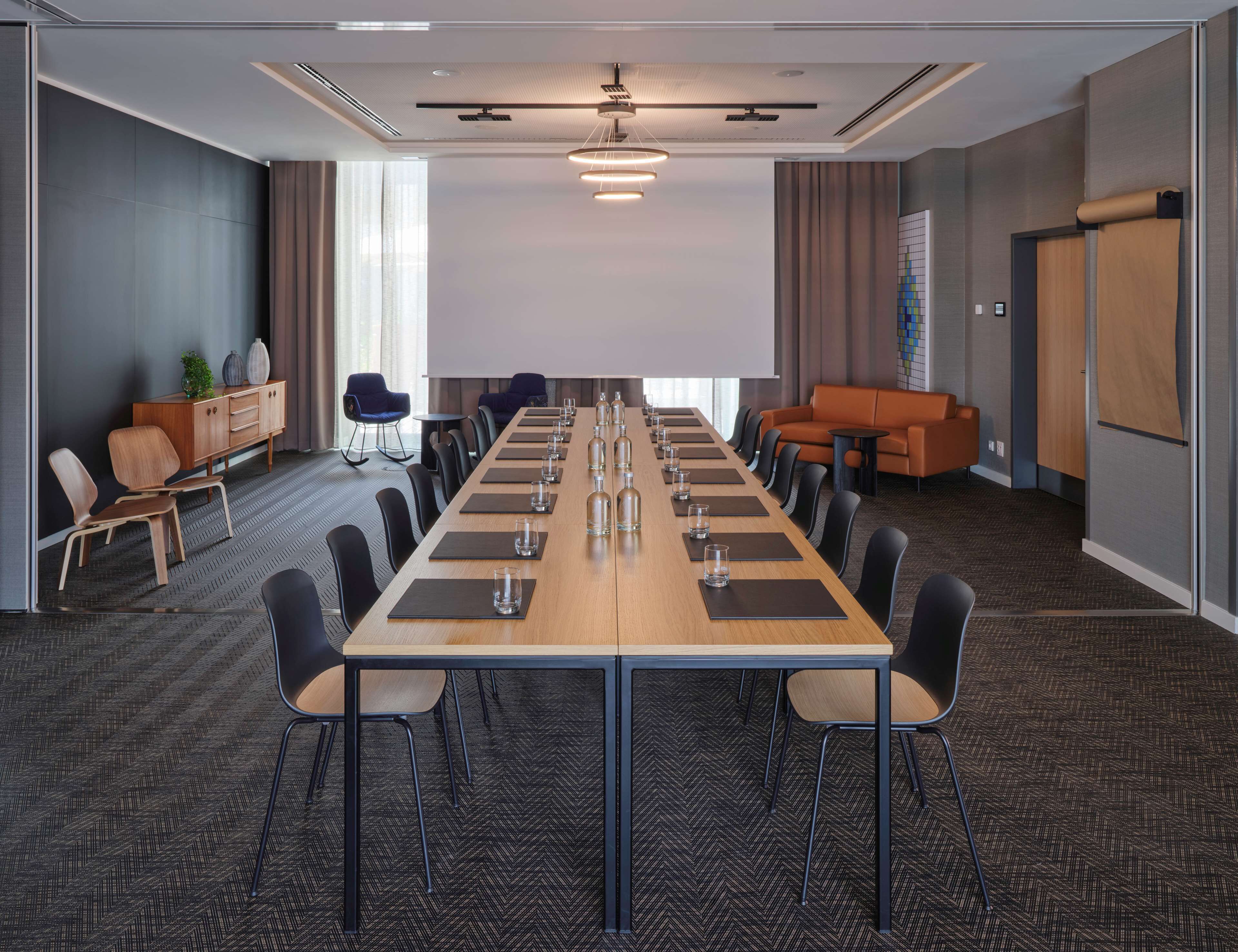 Uma sala de conferências com uma mesa comprida, cadeiras, tela de projetor e um sofá.