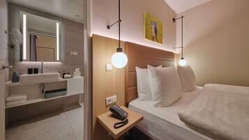 Quarto de hotel moderno com uma cama grande, mesa de cabeceira com telefone, espelho e um pequeno armário com artigos de higiene pessoal.