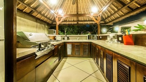Cozinha externa com telhado de palha, churrasqueira embutida e diversos eletrodomésticos.