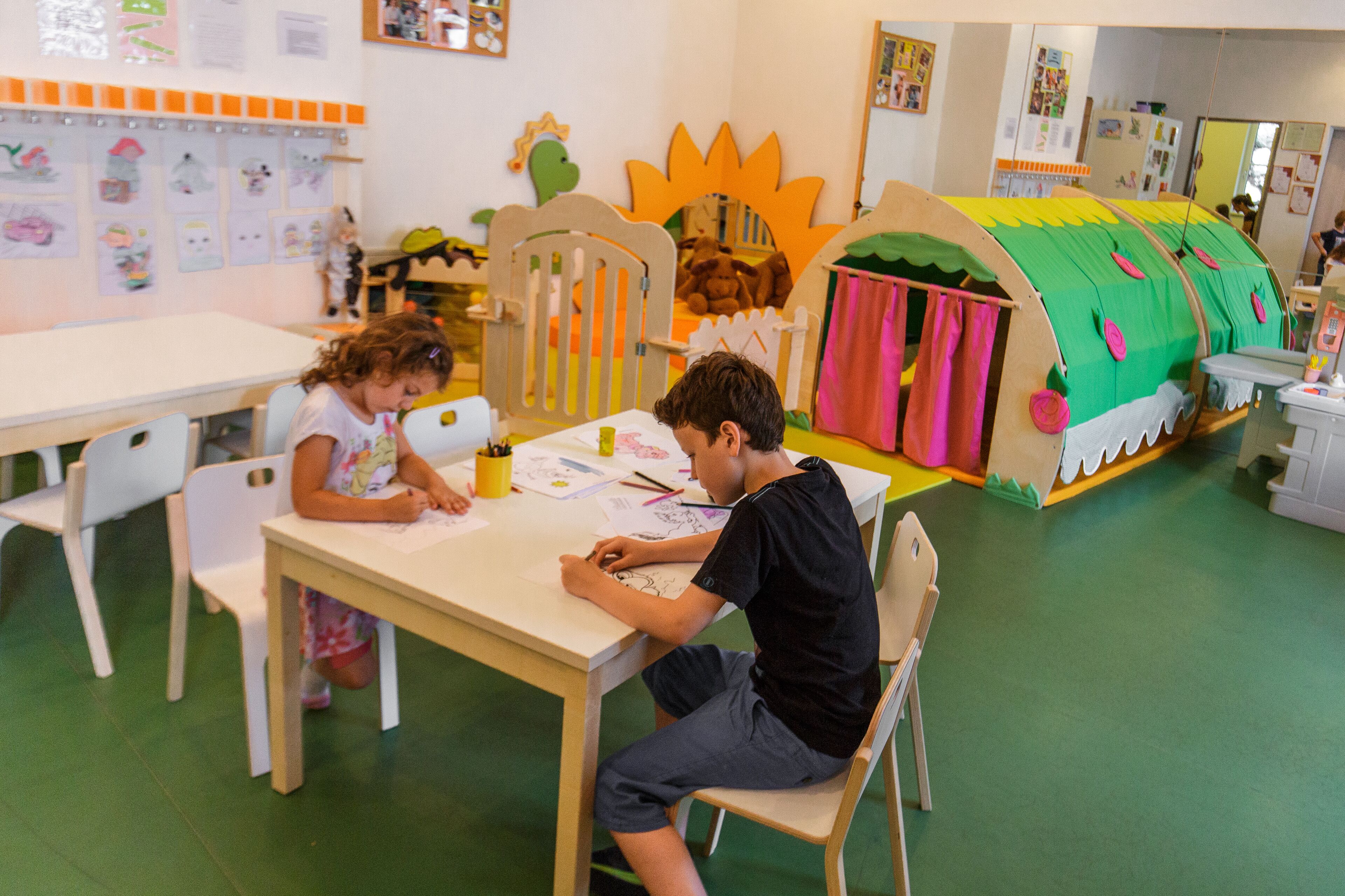 Uma sala de aula com crianças e um adulto à mesa, cercada por cartazes educativos e um espaço de recreação.