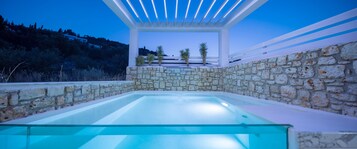 Uma piscina com borda infinita, cercada por vidro, com uma parede de pedra e uma estrutura de pérgula branca.