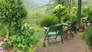 Um alpendre coberto com cadeiras e mesas verdes, rodeado por vegetação exuberante e vista para colinas ao longe.