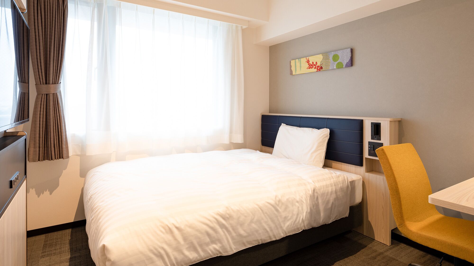 Quarto de hotel com uma cama, uma mesa, uma cadeira, uma TV e uma janela com cortinas.