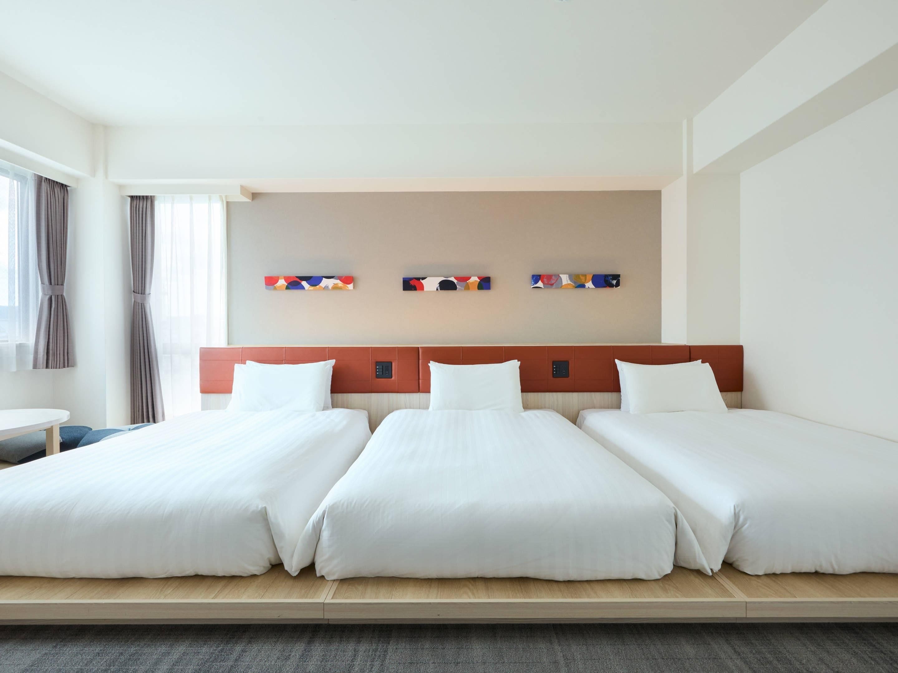 Quarto de hotel com três camas de solteiro, uma mesa redonda pequena e painel de controle fixado na parede.