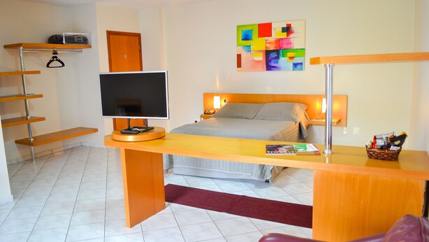 Quarto de hotel com uma cama, uma escrivaninha com televisão, uma cadeira, uma prateleira com uma mala e um quadro abstrato e colorido na parede.