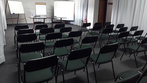 Sala de conferências com fileiras de cadeiras verdes, suportes para quadro branco e uma mesa com garrafas de água.