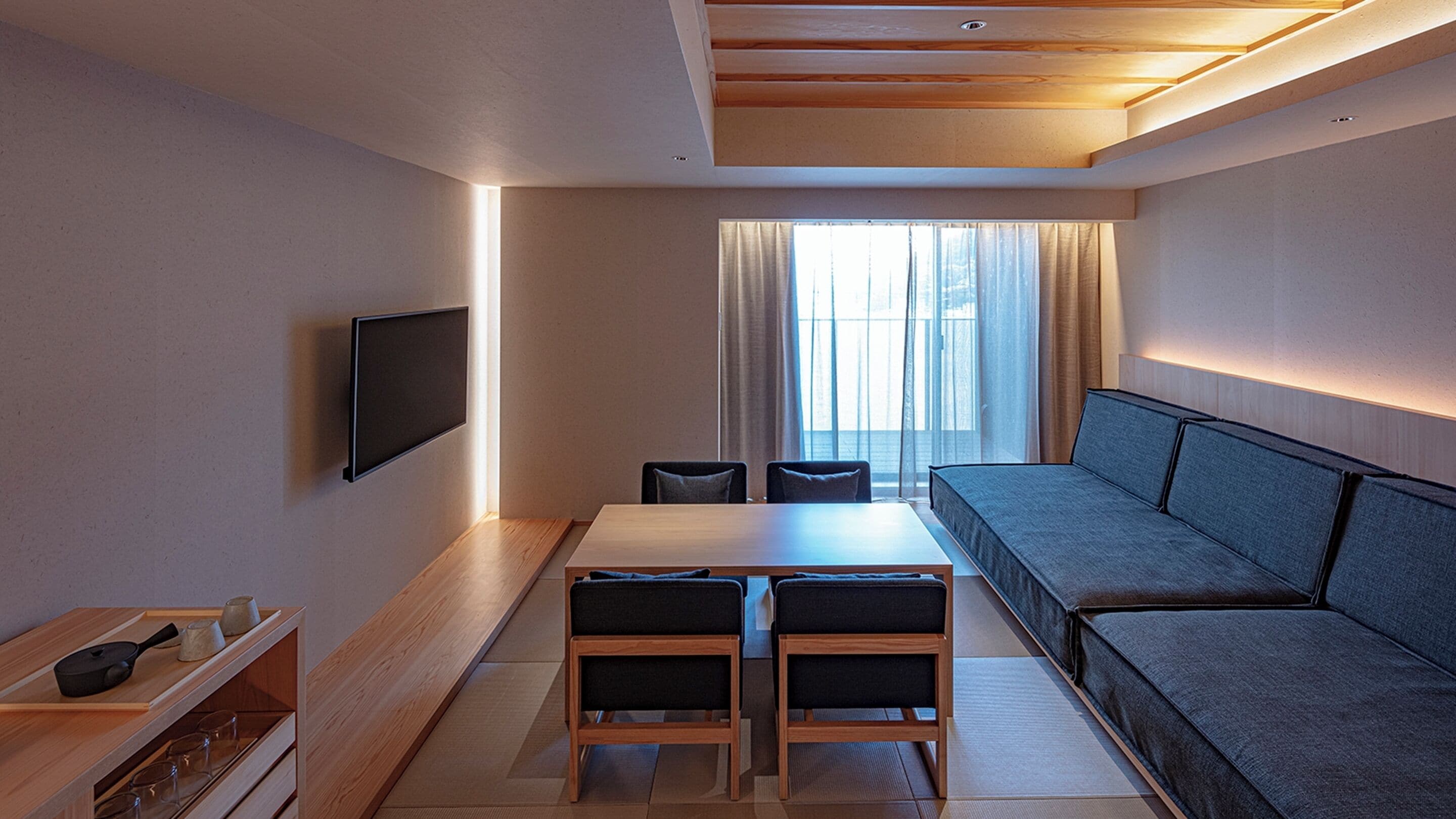 Quarto de hotel moderno com mesa de jantar de madeira, duas cadeiras, um sofá e televisão de tela plana na parede.