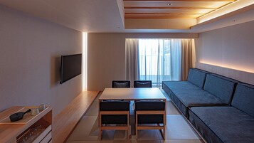 Quarto de hotel moderno com mesa de jantar de madeira, duas cadeiras, um sofá e televisão de tela plana na parede.