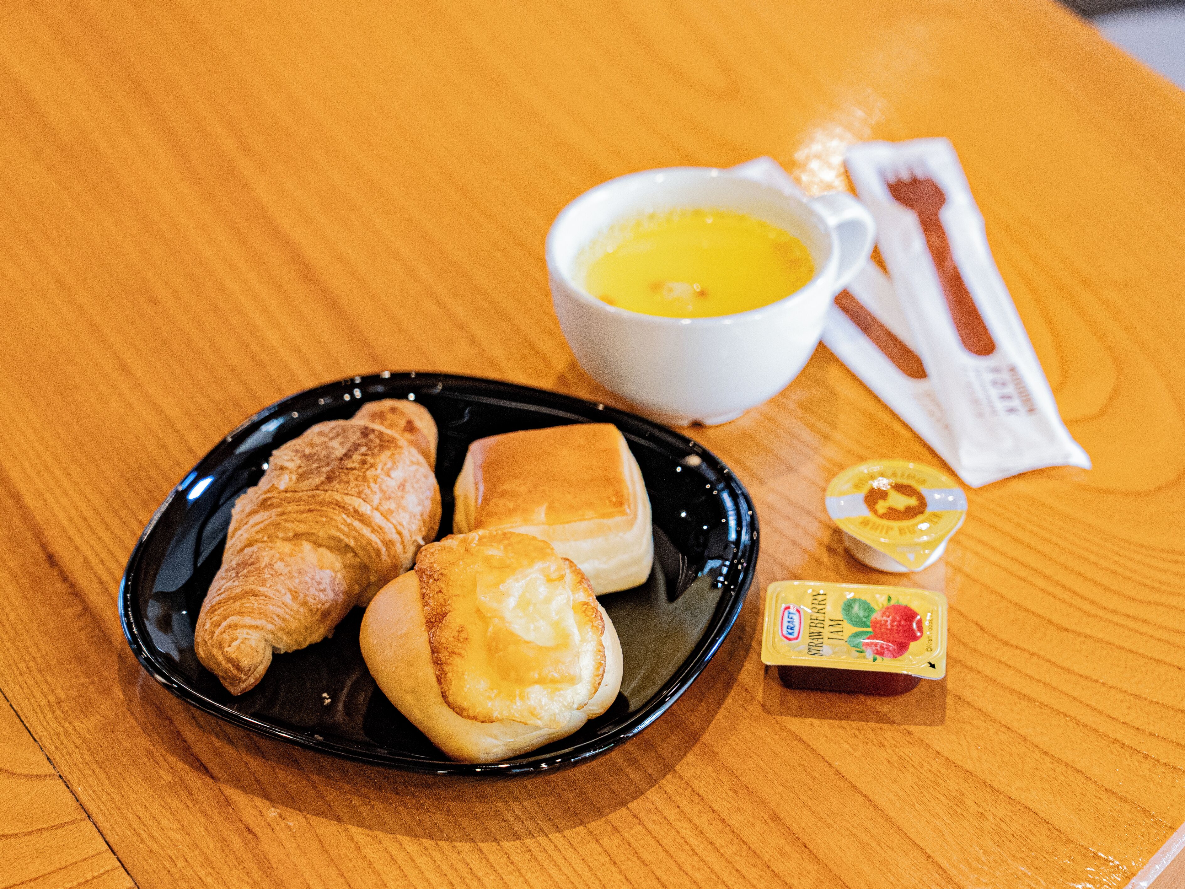 Um prato com croissants, uma tigela de sopa e um pequeno recipiente de geleia sobre uma mesa de madeira.