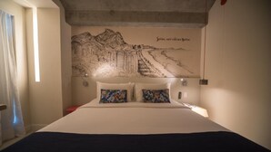 Quarto de hotel com uma cama grande, duas almofadas florais, um mural com vista para a cidade na parede e uma mala vermelha no criado-mudo.