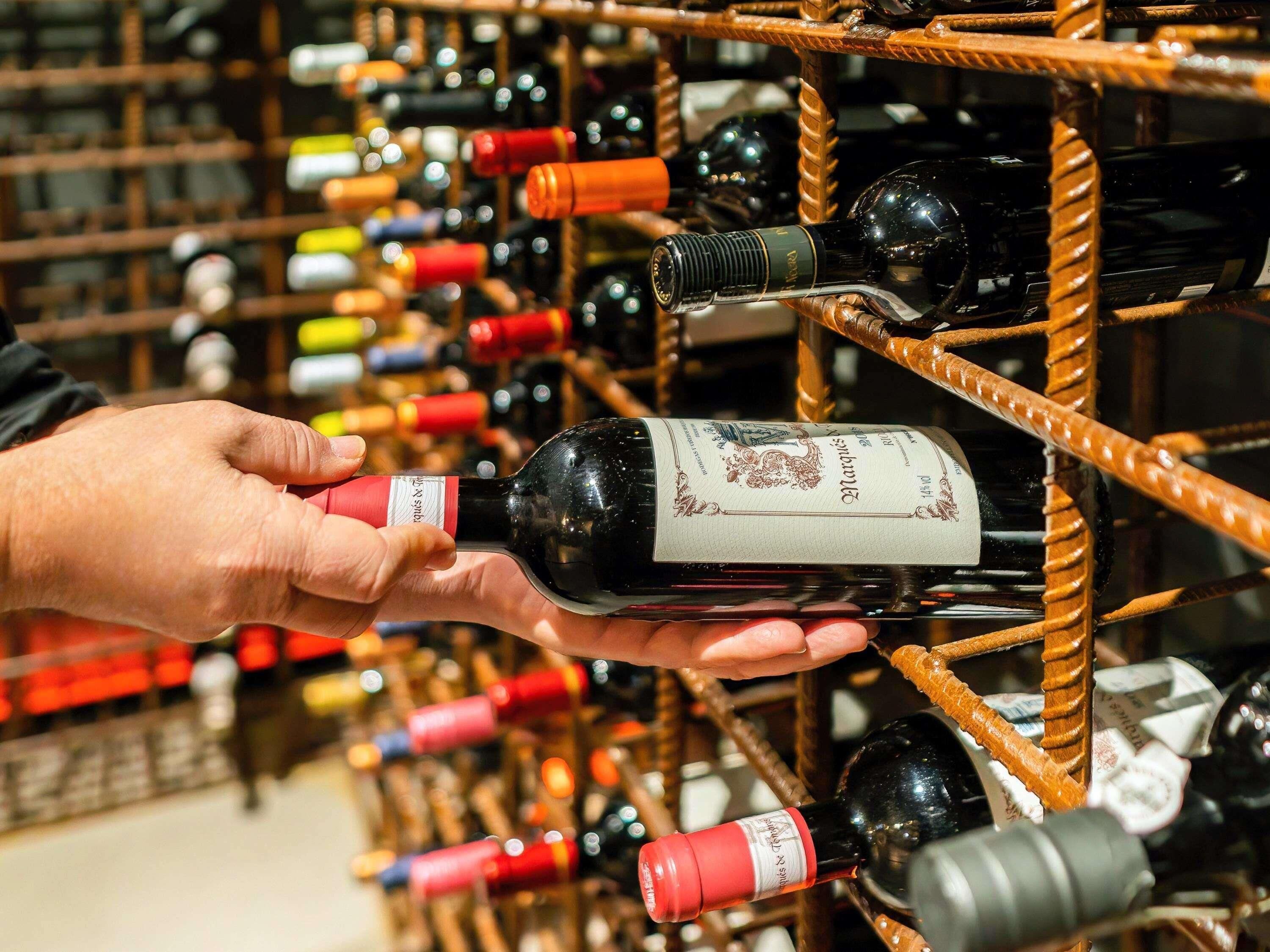 Mão segurando uma garrafa de vinho com etiqueta, rodeada por outras garrafas de vinho em prateleiras.