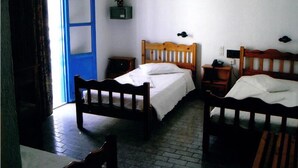 Quarto com duas camas de madeira, uma pequena mesa de cabeceira, uma luminária na parede e uma janela com persianas azuis.