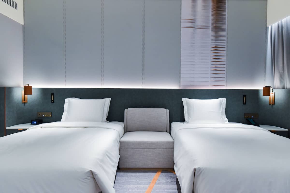 Quarto de hotel com duas camas, um espaço de estar pequeno e uma janela grande com persianas.