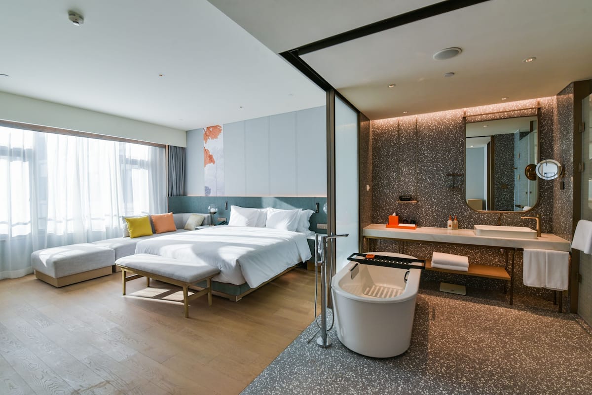 Quarto de hotel moderno com uma cama grande, área de estar, banheira e banheiro com espelho e pia.