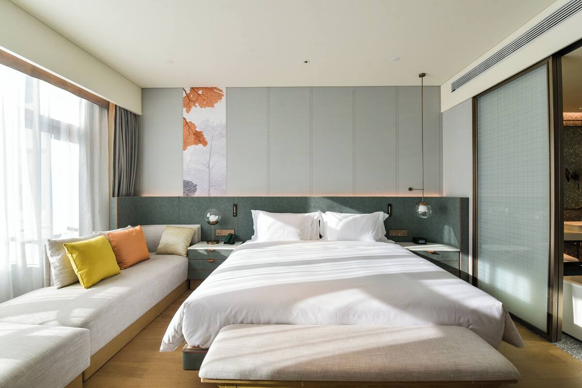 Quarto de hotel moderno com uma cama grande, um sofá-cama, uma mesinha de cabeceira e uma janela ampla com cortinas.