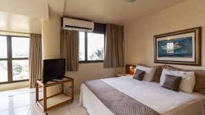 Quarto de hotel com uma cama, uma televisão sobre suporte de madeira, um aparelho de ar condicionado e um quadro na parede.
