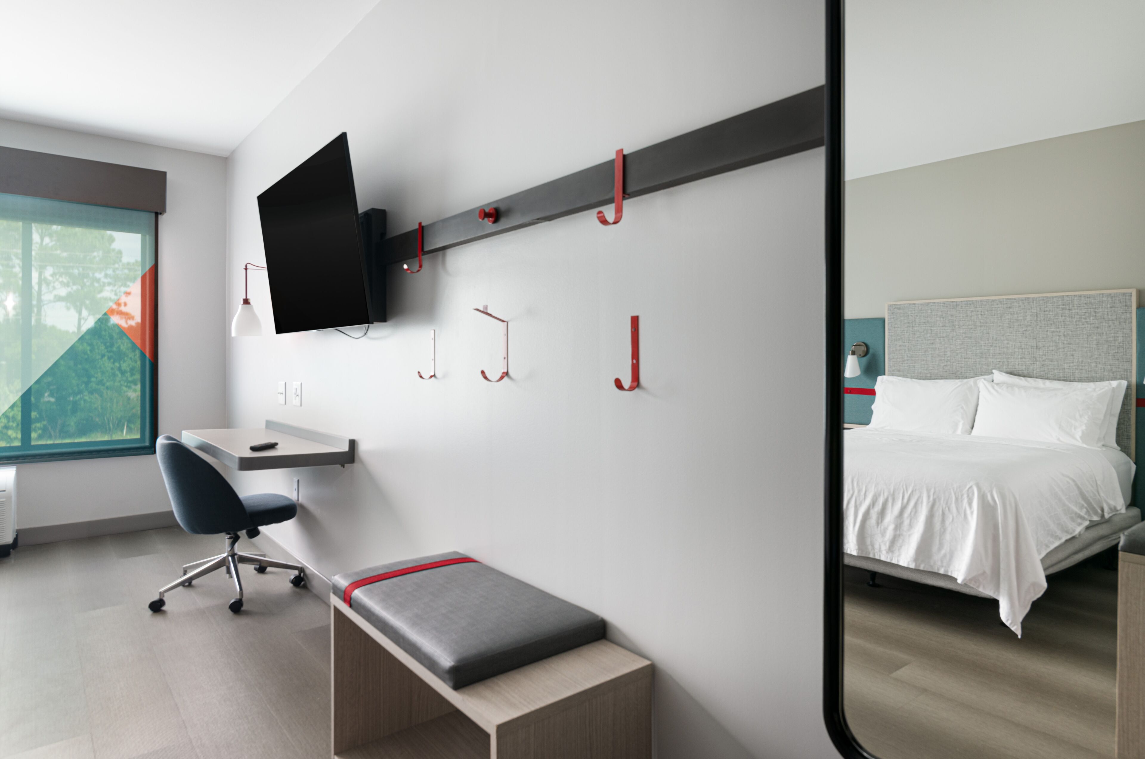 Quarto de hotel moderno com cama, escrivaninha com cadeira, um banco, televisão presa à parede e janela com vista para árvores.