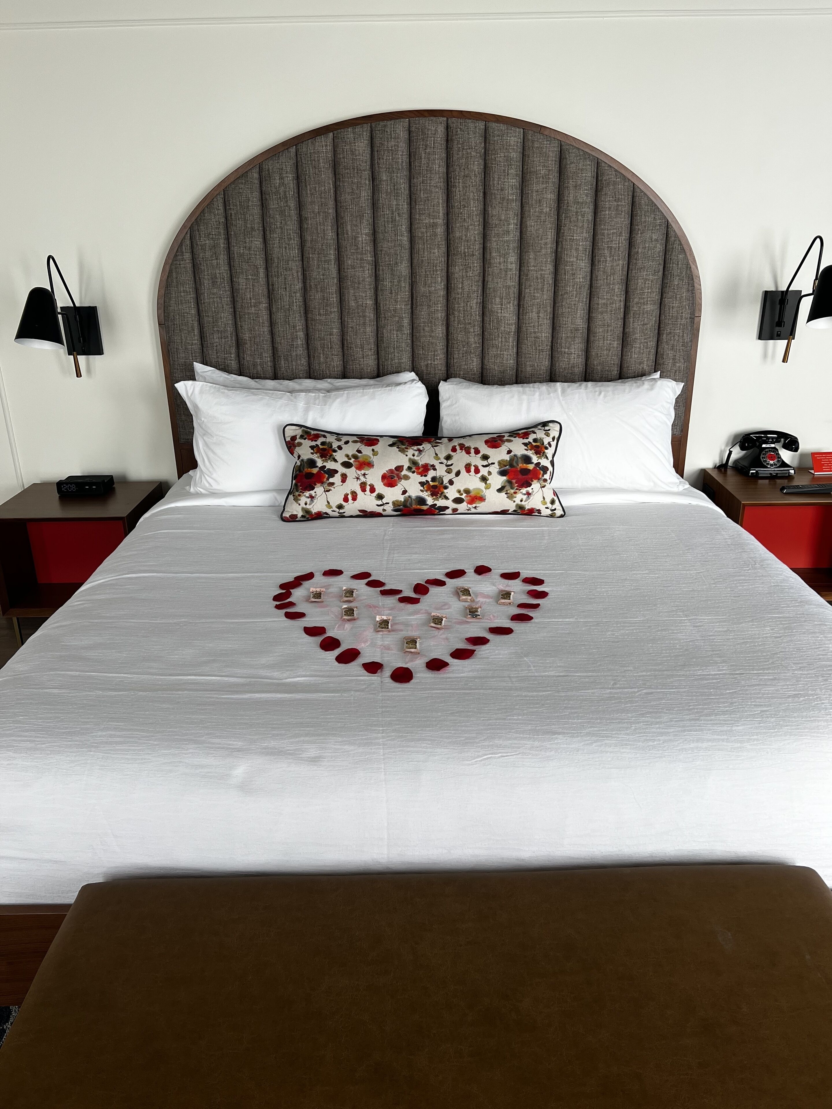 Uma cama bem arrumada com uma decoração em formato de coração na colcha, uma cabeceira de madeira e duas mesas de cabeceira com abajures.