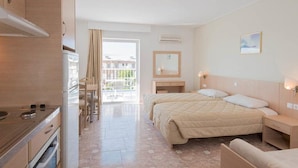 Quarto de hotel com cozinha compacta, uma cama com colcha bege, mesa de cabeceira, espelho e vista para o exterior.