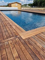Um deck de madeira rodeia uma piscina azul cristalina, com uma cabana de madeira ao fundo.