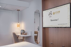Quarto de hotel com uma cama, uma mesinha com chaleira, uma cadeira e uma televisão na parede exibindo uma mensagem de boas-vindas.