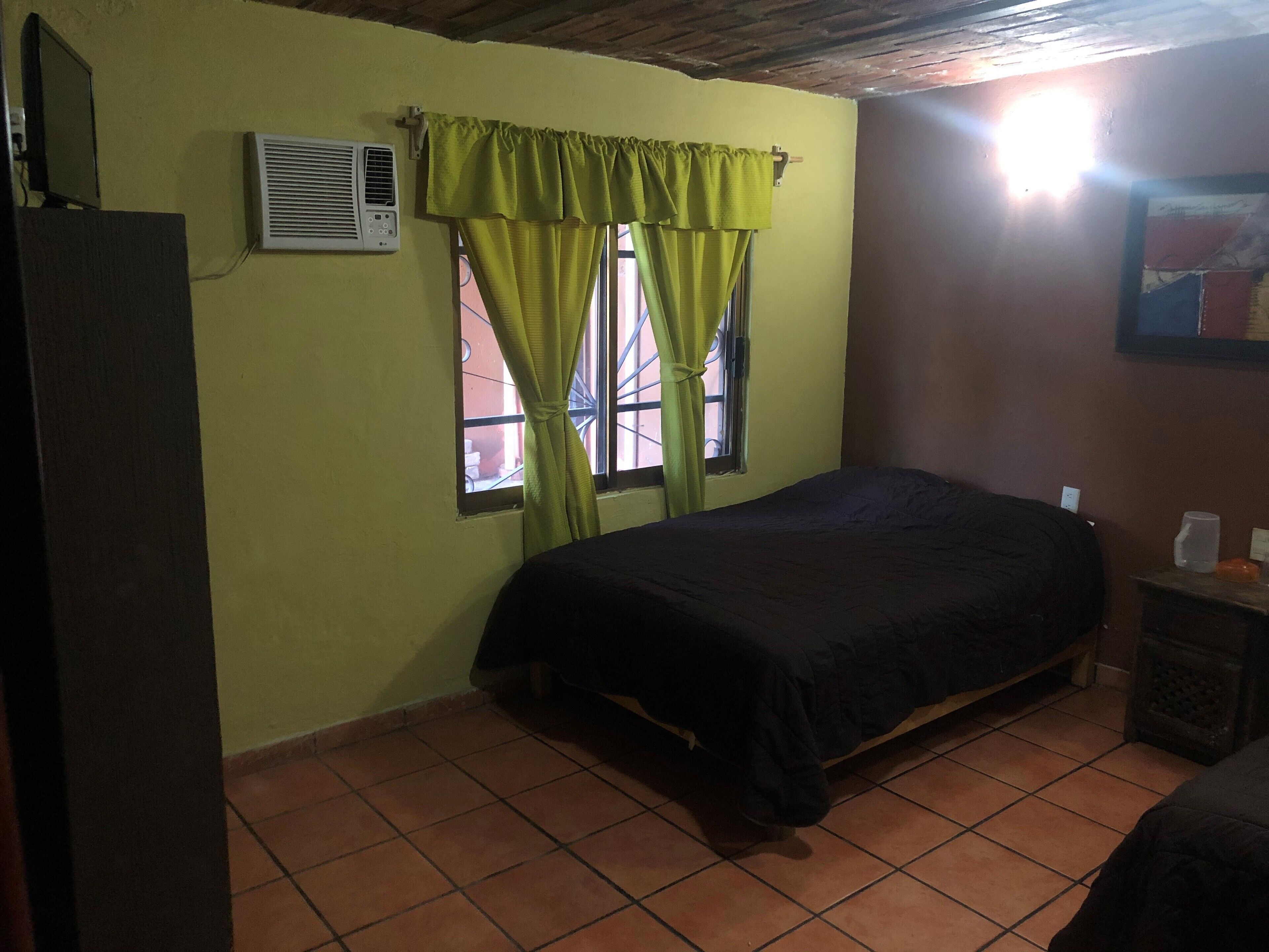 Quarto com cama, janela com cortinas, aparelho de ar condicionado e piso ladrilhado.
