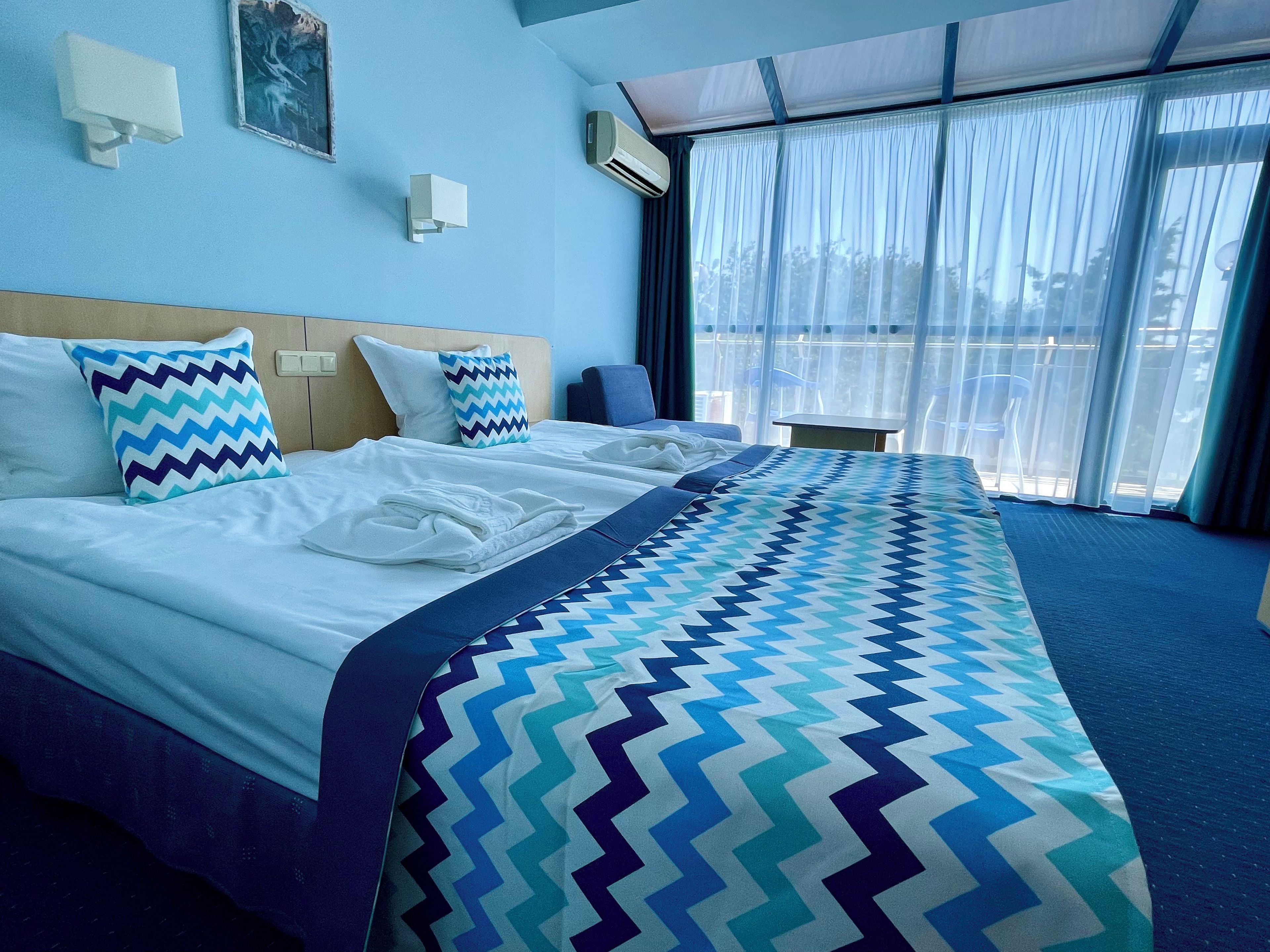 Quarto de hotel com uma cama grande, uma mesinha e vista para o exterior através de grandes janelas.