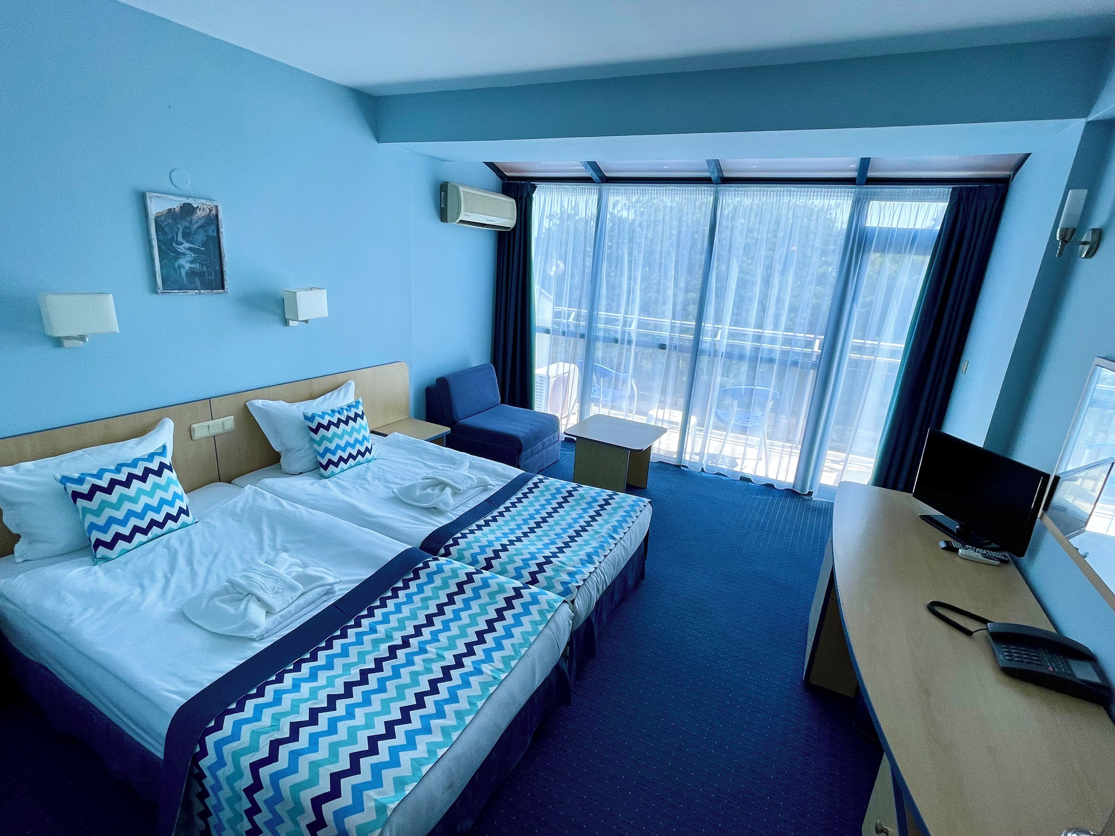 Quarto de hotel com cama, escrivaninha, cadeira, televisão e janela com cortinas.