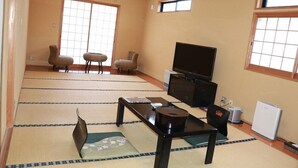 Uma sala de estar tradicional japonesa com tatames, uma televisão, uma pequena mesa e um sofá.