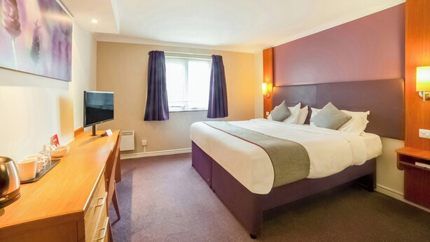 Quarto de hotel com cama, escrivaninha com computador, televisão e janela com cortinas.